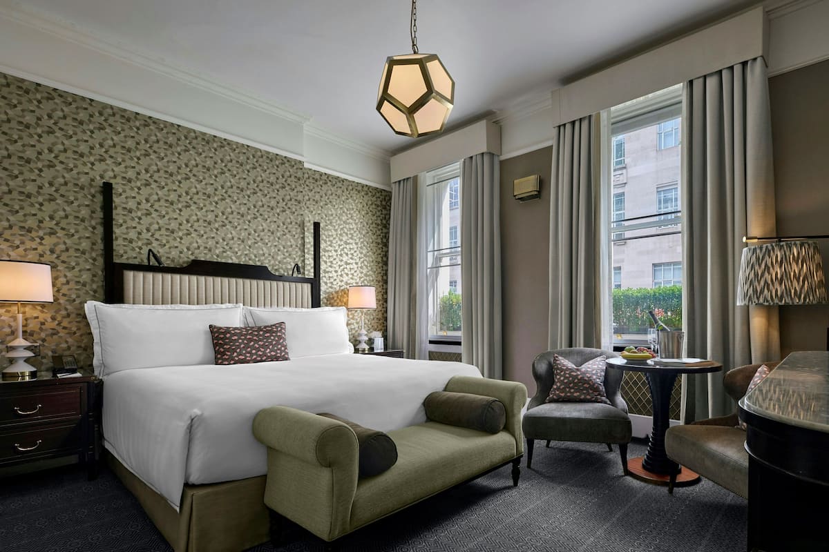 Quarto de hotel com uma cama grande, uma poltrona pequena, uma mesa de apoio e vista para o exterior através de uma janela.