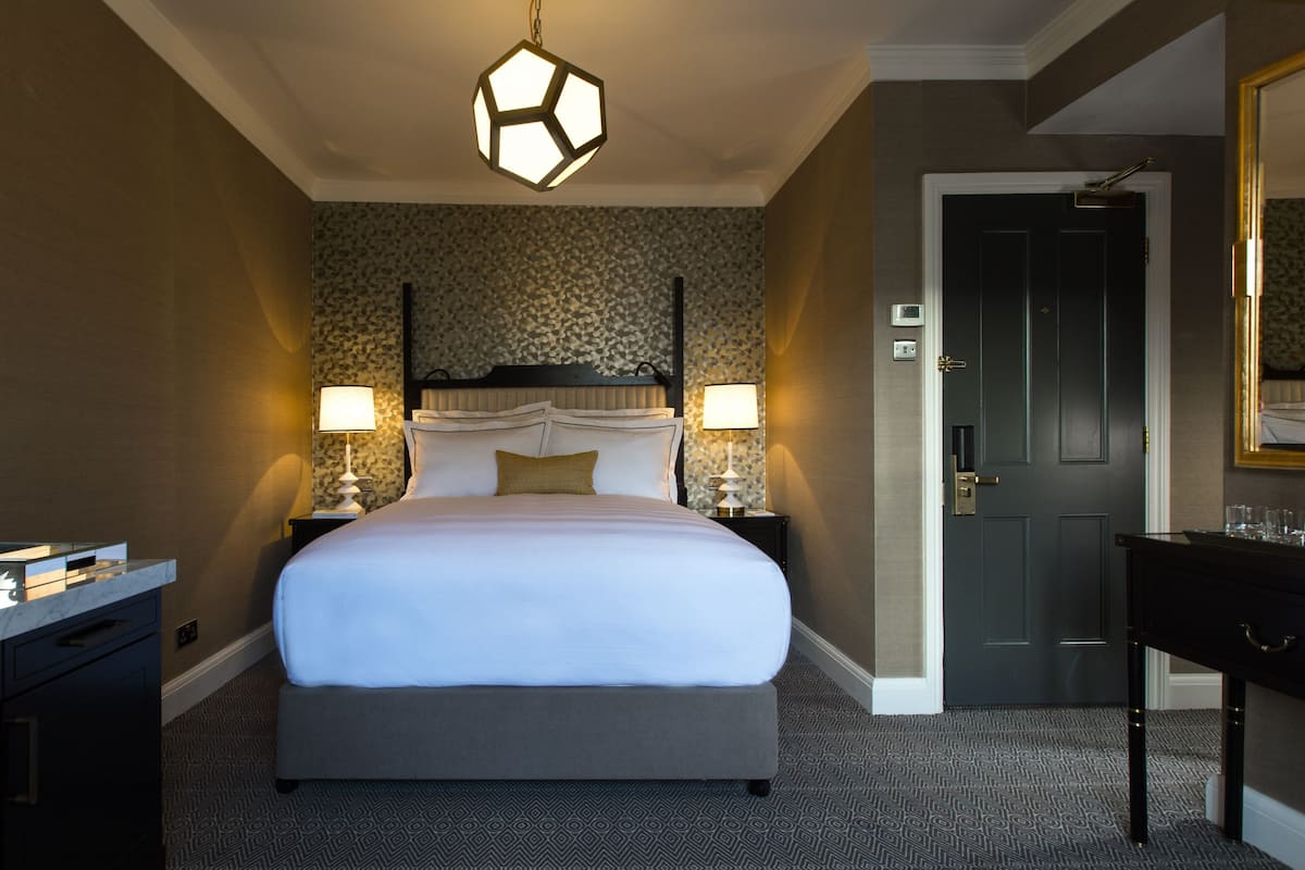 Quarto de hotel com uma cama grande, duas luminárias de cabeceira, um tapete com padrão geométrico, uma porta escura e um espelho na parede.