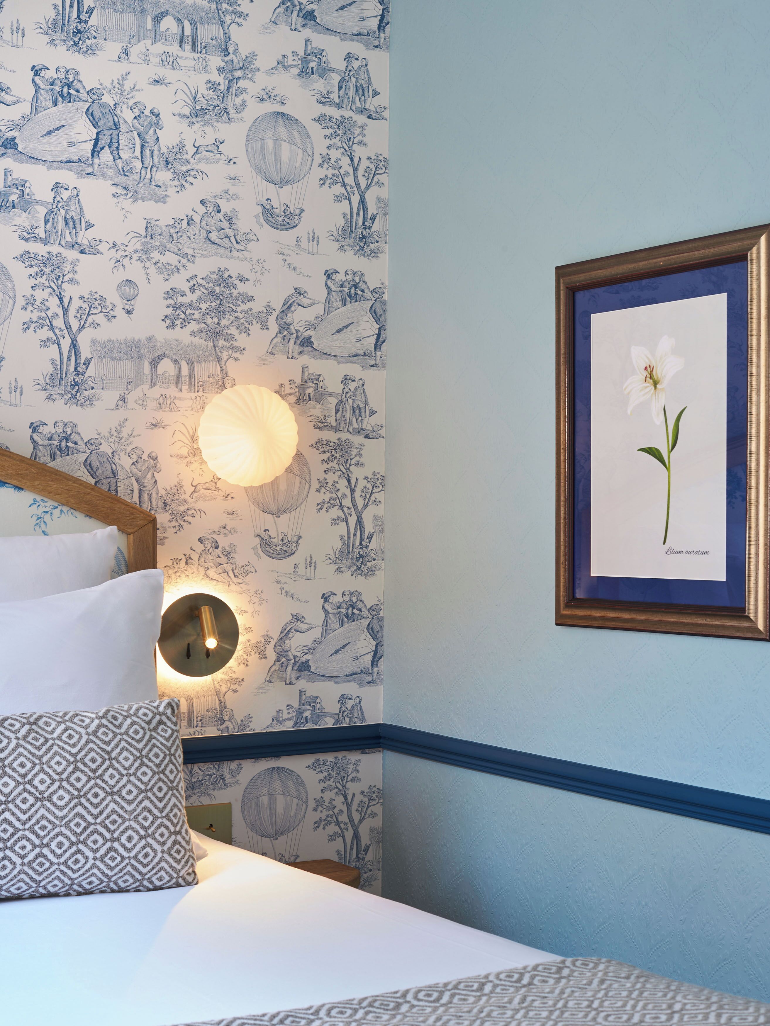 Quarto com cama, almofadas, luminária na parede e gravura botânica emoldurada.