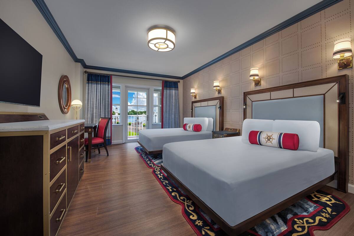 Quarto de hotel com duas camas, uma escrivaninha, uma cadeira e uma janela grande com cortinas.