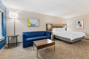 Quarto de hotel moderno com sofá azul, mesa de centro de madeira, cama com roupa branca, mesa de cabeceira, abajur e arte abstrata na parede.