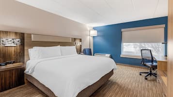 Quarto de hotel com uma cama grande, uma escrivaninha com cadeira, mesa de cabeceira, televisão fixada na parede e janela com cortinas.