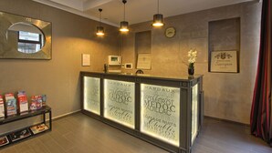 Uma área de recepção com design moderno, destacando um balcão com painéis de vidro iluminados exibindo texto, iluminação pendente, um espelho grande, um relógio e um certificado emoldurado na parede.