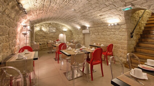 Uma sala de jantar com paredes de pedra, mesas e cadeiras, uma escada e vista para outro ambiente.