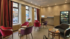 Um saguão de hotel com cadeiras vermelhas e roxas, uma máquina de café e vista para a rua pela janela.