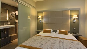 Quarto de hotel com uma cama grande, duas mesinhas de cabeceira, um criado-mudo, um espelho, banheiro com pia e porta para toalhas, e uma porta de correr.