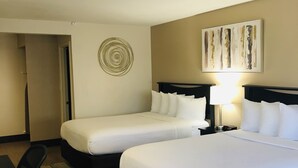 Quarto de hotel com duas camas de solteiro e uma cama maior, uma escrivaninha, uma cadeira, um abajur e uma obra de arte na parede.
