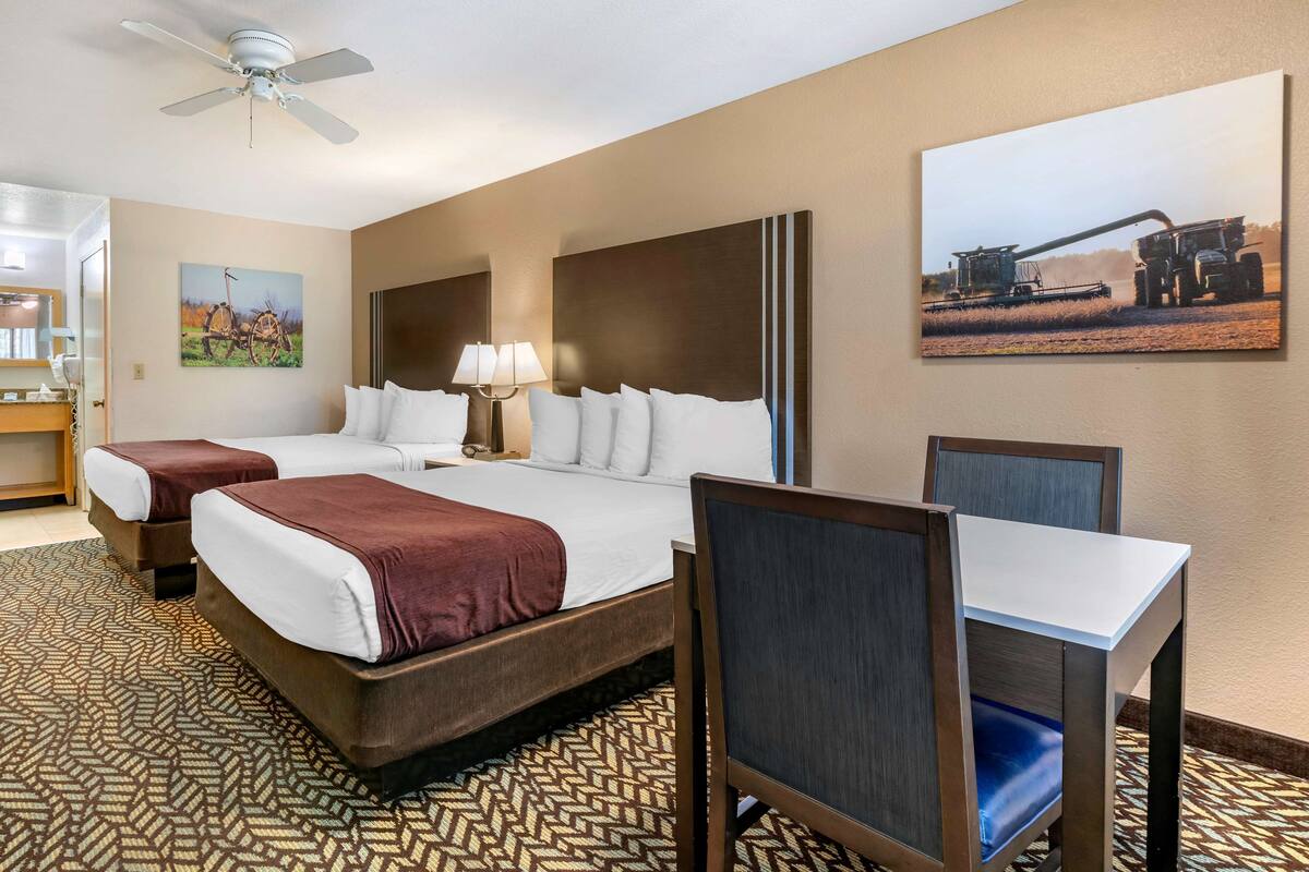 Quarto de hotel com duas camas, uma escrivaninha, uma cadeira, ventoinha no teto, abajur e um quadro na parede.