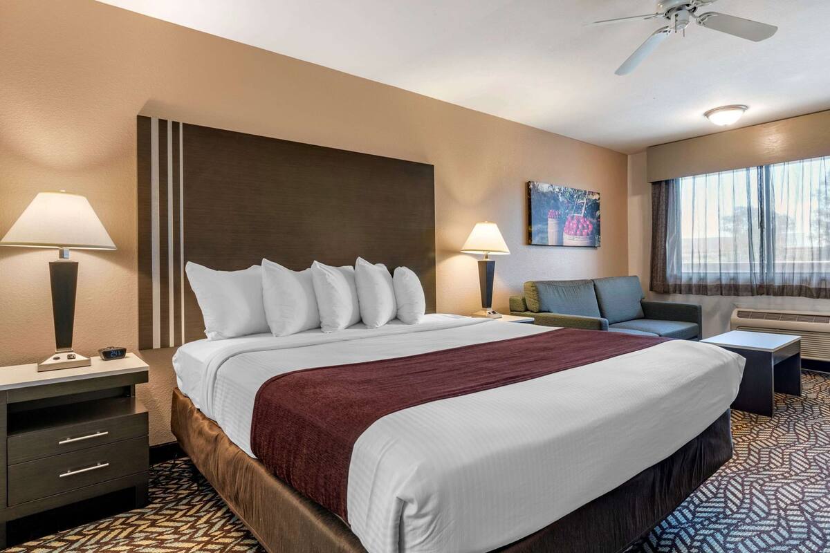 Quarto de hotel com uma cama grande, duas mesas de cabeceira, uma escrivaninha, uma cadeira, um ventilador de teto e uma janela com cortinas.