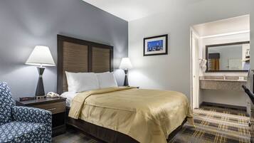 Quarto de hotel com cama, duas luminárias de cabeceira, uma cadeira, mesa de cabeceira, espelho e um quadro na parede.