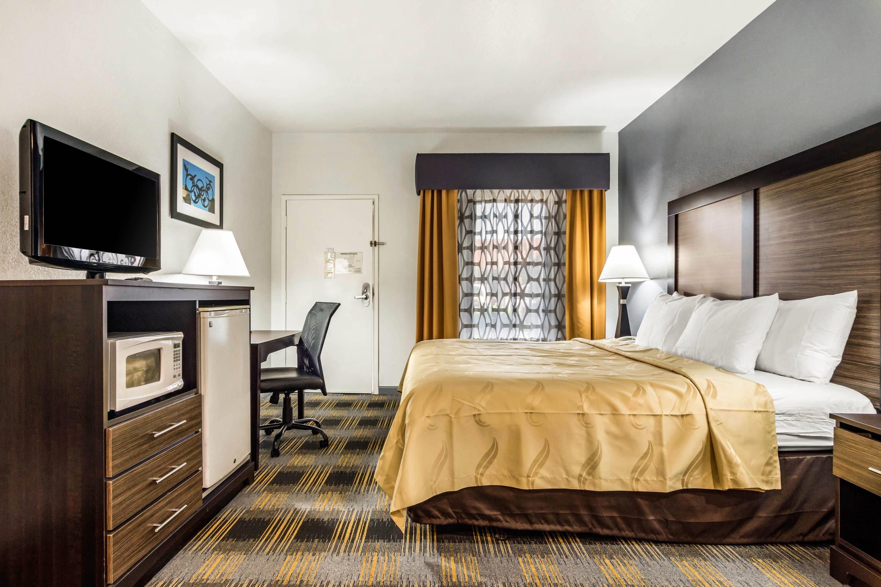 Quarto de hotel com cama, escrivaninha, televisão, frigorífico e janela com cortinas.