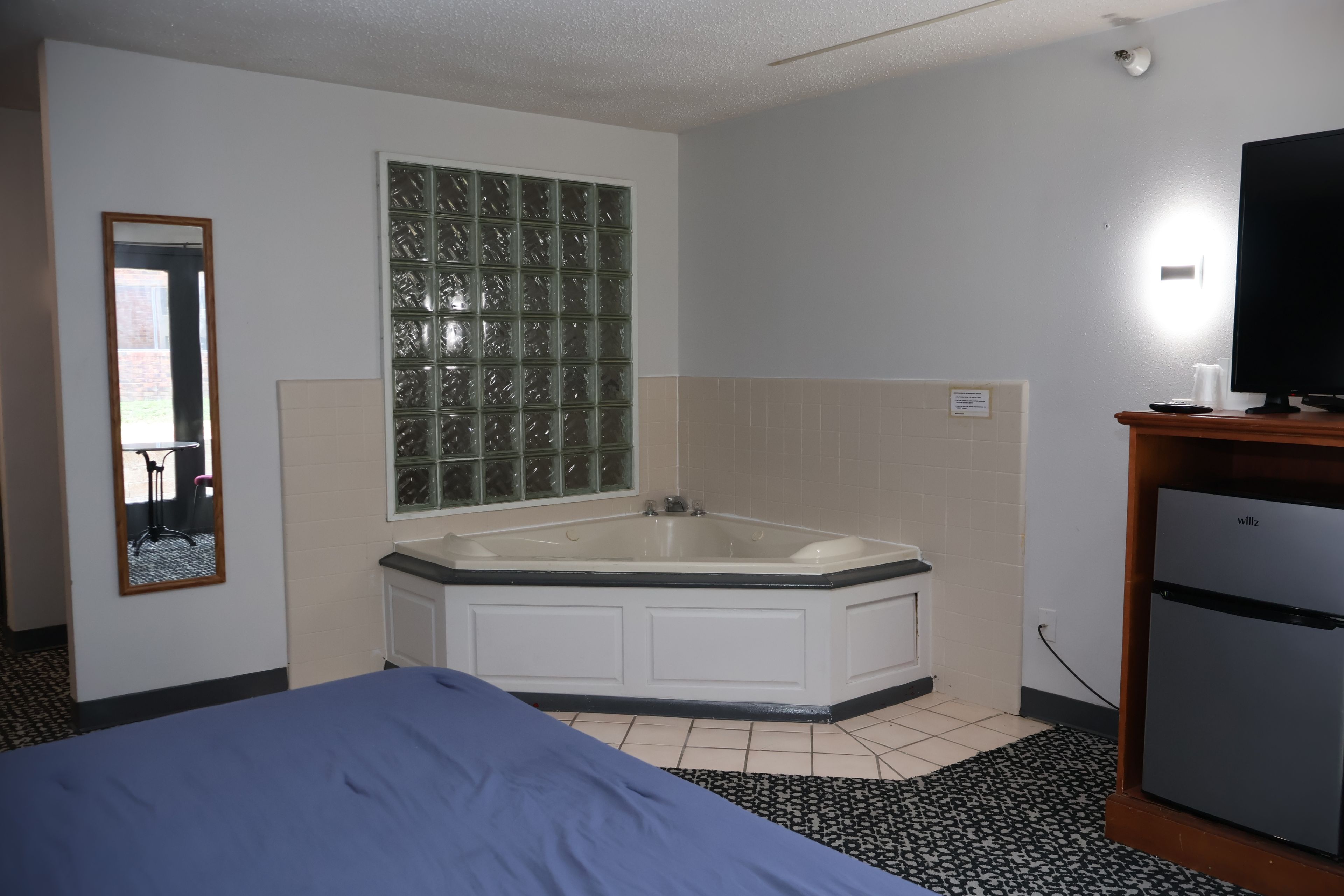 Quarto com uma banheira grande, cama com cobertura azul, um espelho e televisão sobre suporte.