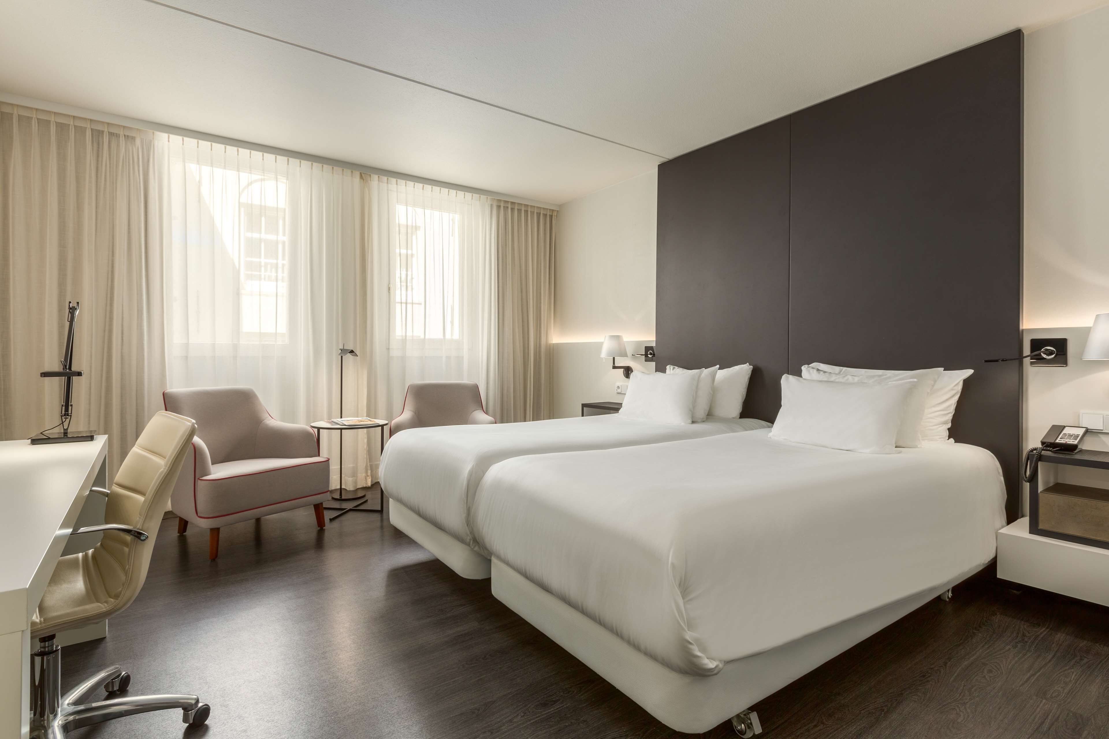 Quarto de hotel com duas camas, uma escrivaninha, uma cadeira, uma mesinha e um abajur.