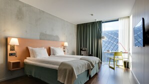 Quarto de hotel moderno com uma cama grande, duas mesas de cabeceira com abajures, uma escrivaninha com cadeira, uma TV na parede e vista para o céu pela janela.