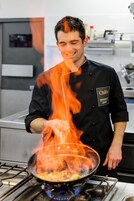 Um cozinheiro com uniforme preto e etiqueta de identificação, cozinhando com chamas visíveis em uma panela no fogão.