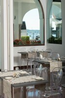 Restaurante com cadeiras e mesas de vidro transparente, vista da cidade através de grandes janelas e um espelho refletindo a paisagem externa.