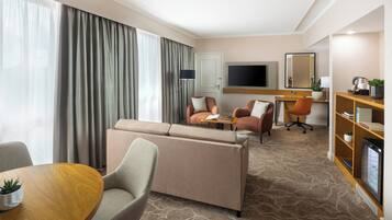 Quarto de hotel com sofá, poltronas, secretária, televisão e estante de livros.
