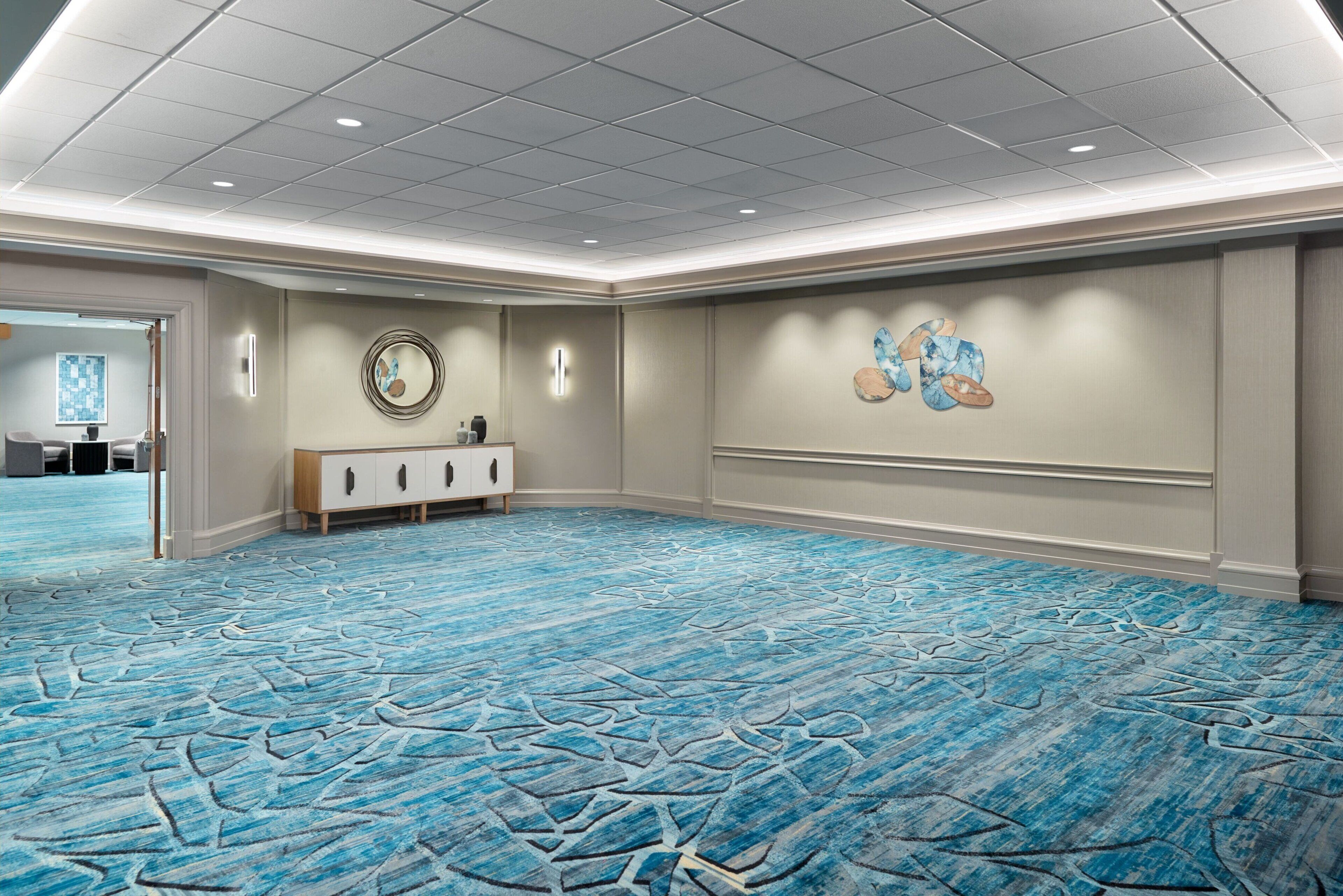 Uma sala de conferências espaçosa, com carpete estampado em azul, um armário branco e uma parede com espelho circular e obras de arte.