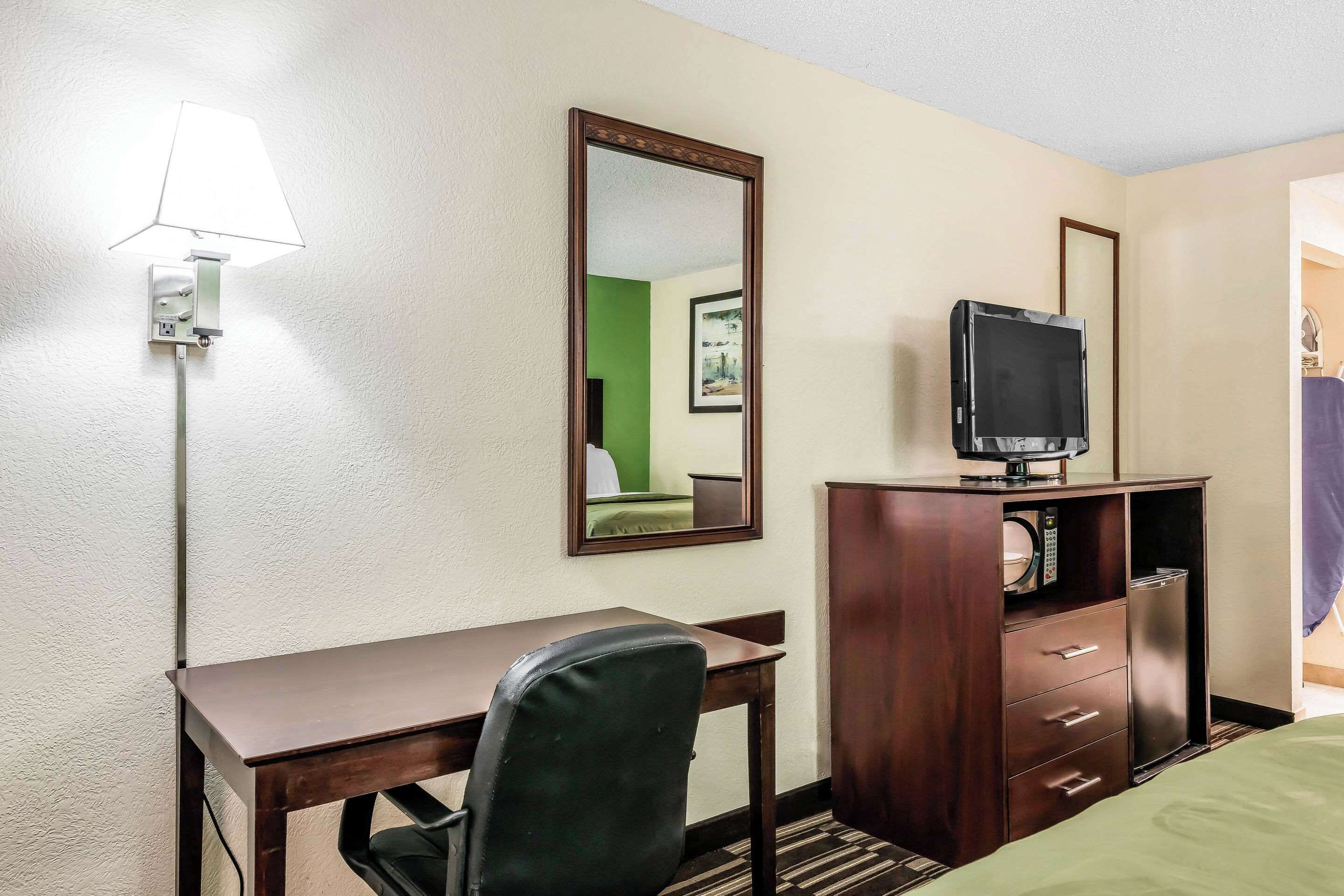 Quarto de hotel com escrivaninha, cadeira, televisão, cômoda e espelho.