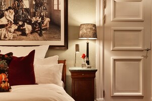 Quarto com cama, almofadas, mesa de cabeceira com luminária, um vaso com flor e um quadro na parede.