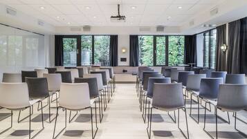 Sala de conferências com fileiras de cadeiras, um projetor e grandes janelas com cortinas.
