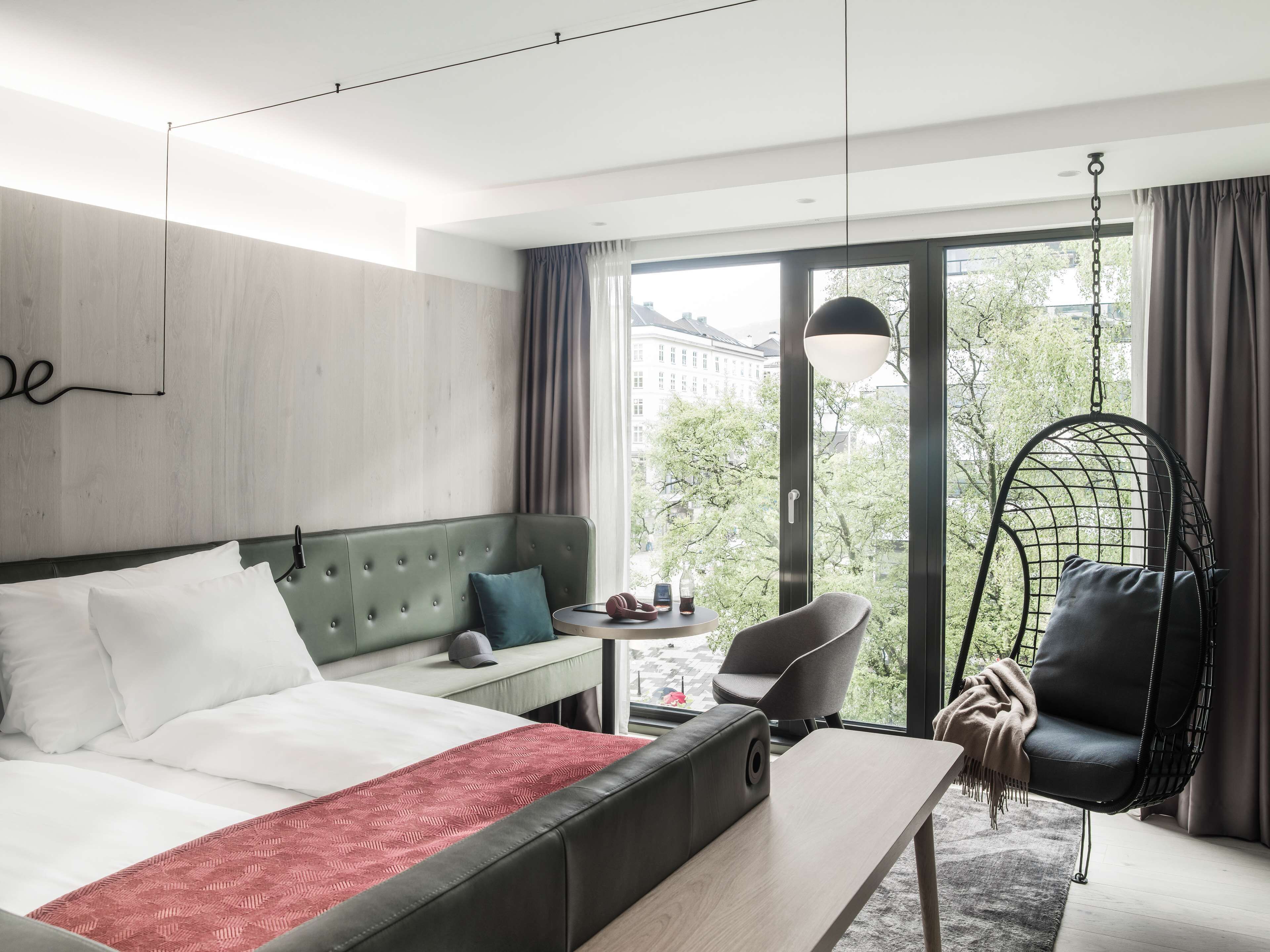 Quarto de hotel moderno com cama, sofá, cadeira suspensa e vista da cidade através de grandes janelas.