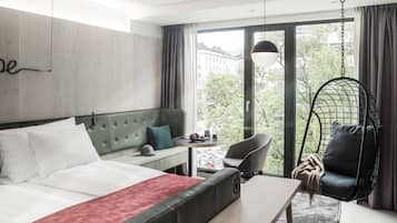 Quarto de hotel moderno com cama, sofá, cadeira suspensa e vista da cidade através de grandes janelas.