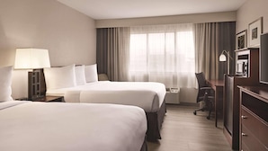 Quarto de hotel com duas camas, uma escrivaninha, uma cadeira, uma televisão e um abajur.