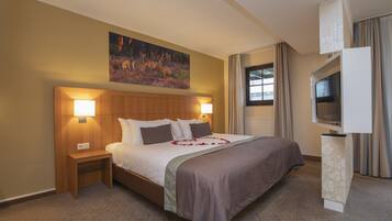 Quarto de hotel com uma cama grande, duas mesas de cabeceira, uma televisão de tela plana, um quadro na parede e uma janela com cortinas.