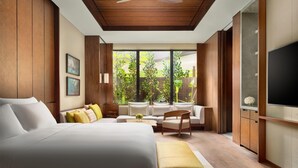 Quarto moderno com uma cama grande, área de estar com cadeiras, televisão e vista para o verde lá fora.