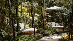 Um jardim interior exuberante, com um caminho que leva a uma área de estar com móveis de vime e almofadas brancas.