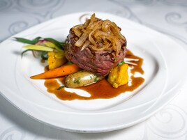 Prato servido com carne bovina coberta por cebolas caramelizadas, acompanhada de vegetais e molho.