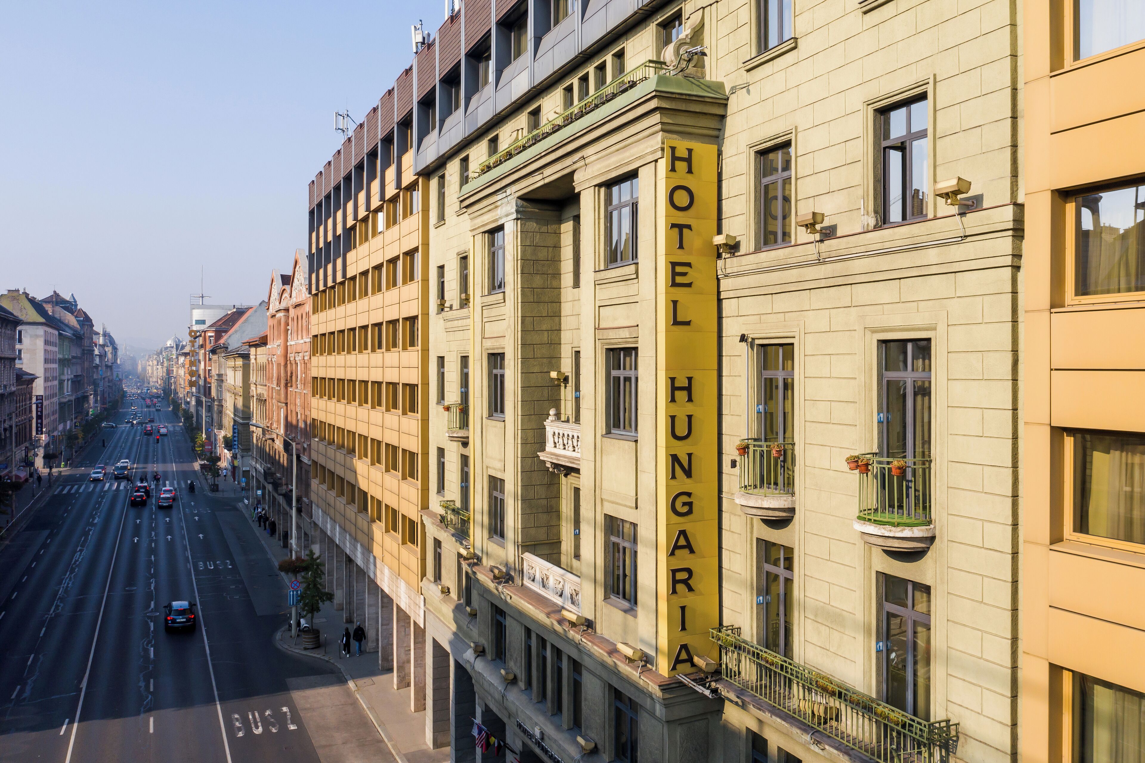 Um hotel de vários andares com uma placa grande de "HOTEL HUNGARIA" na fachada. O edifício apresenta janelas amplas e varandas com plantas em vasos.