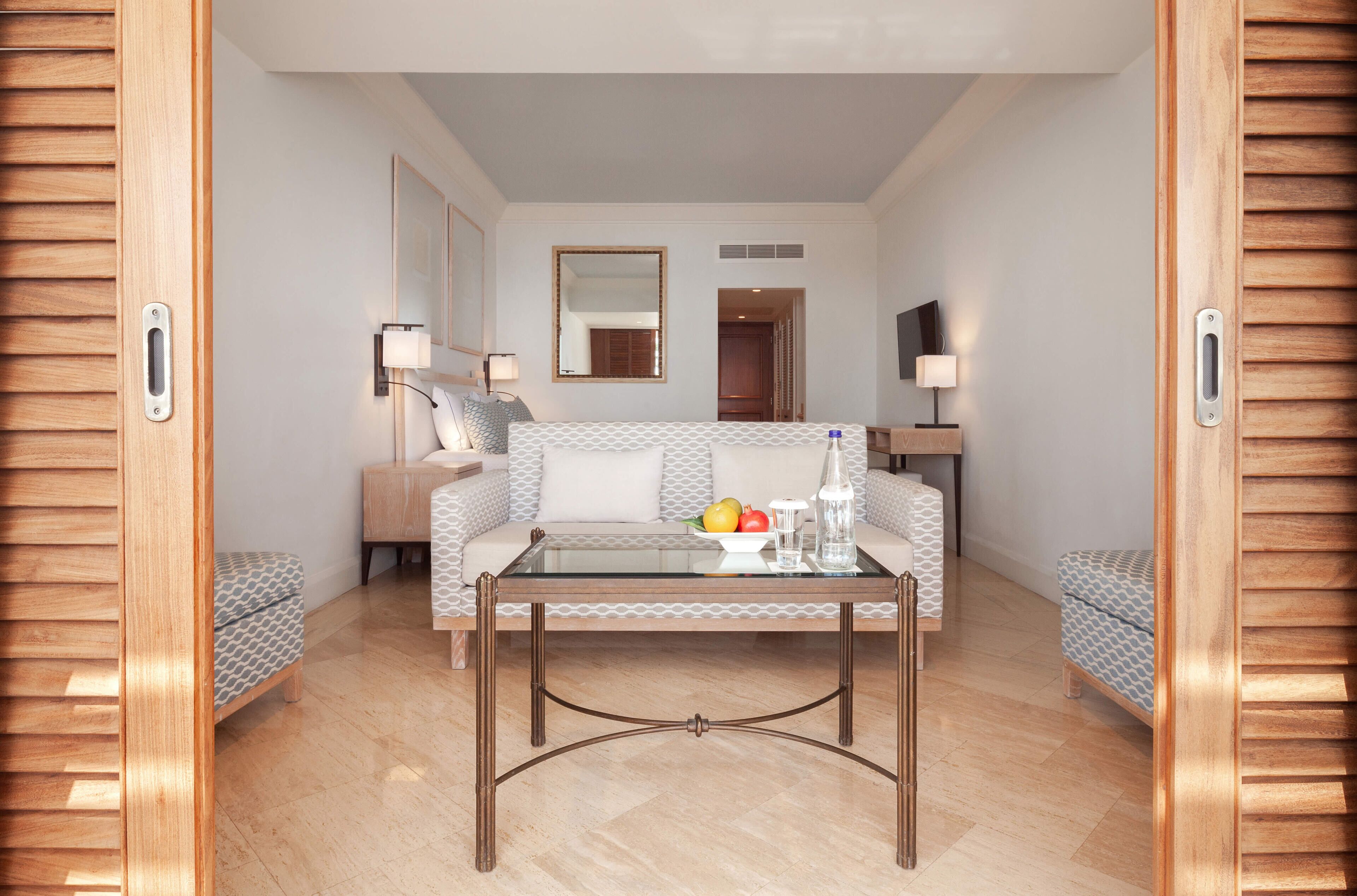 Quarto de hotel moderno com mesa de centro de vidro, sofá estampado e piso de madeira.