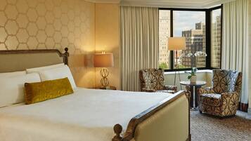 Quarto de hotel com uma cama grande, duas cadeiras, uma mesa pequena e vista para a cidade através de janelas amplas.