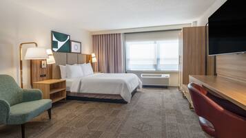 Quarto de hotel com cama, escrivaninha, cadeira, televisão, luminária e janela com cortinas.