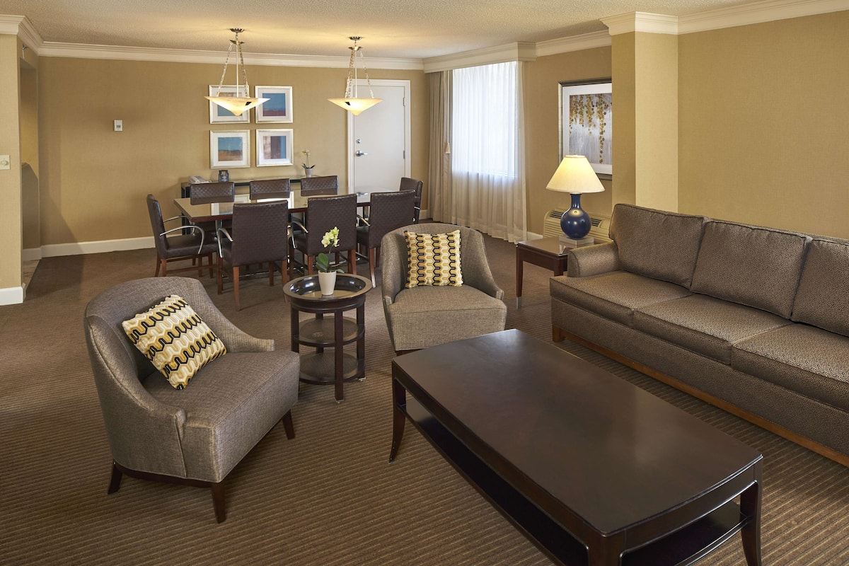 Uma sala de hotel com sofá, poltronas, mesa de centro, área de jantar com cadeiras e uma mesa, e um candelabro.