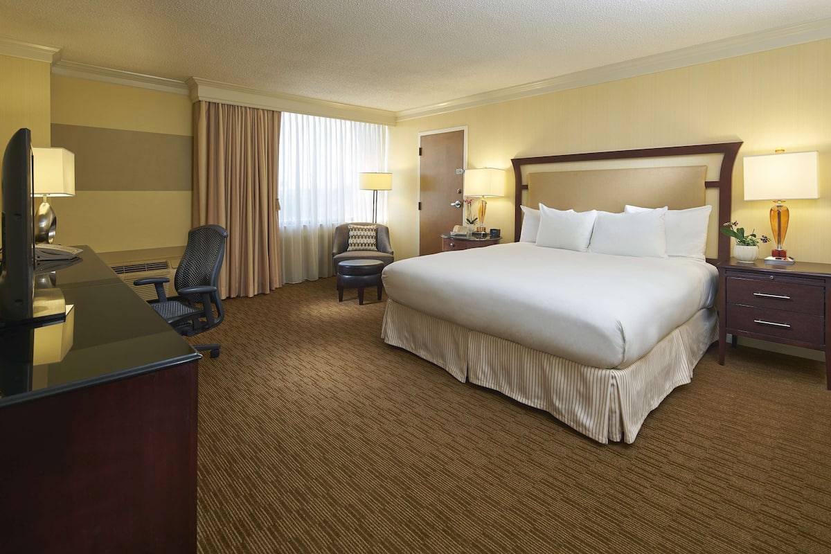 Quarto de hotel com uma cama grande, uma escrivaninha com computador, uma cadeira e mesa de cabeceira com luminárias.