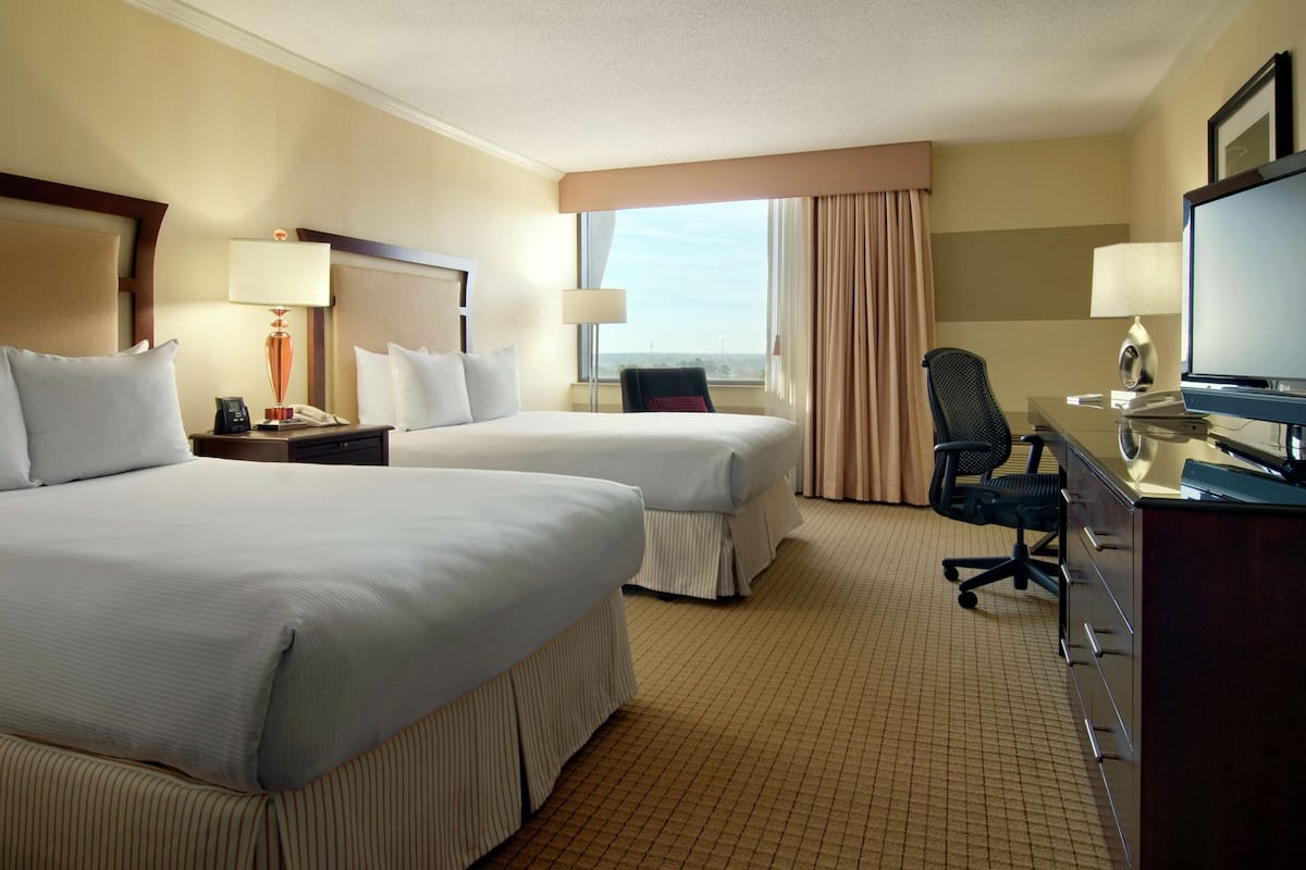 Quarto de hotel com duas camas, uma escrivaninha, uma cadeira, uma televisão e uma janela com cortinas.