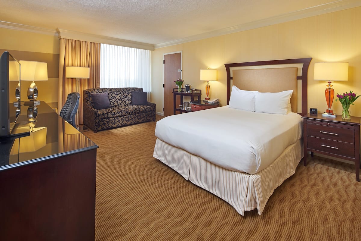 Quarto de hotel com uma cama grande, uma escrivaninha com espelho, uma cadeira, mesa de cabeceira com abajur e um vaso com flores.