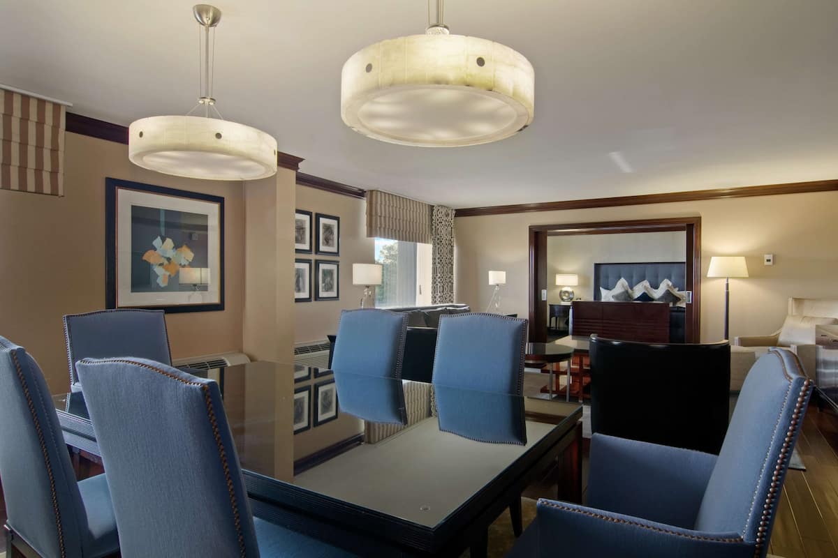 Quarto de hotel com área de jantar, mesa de vidro e cadeiras estofadas em azul, um espelho grande e um lustre.