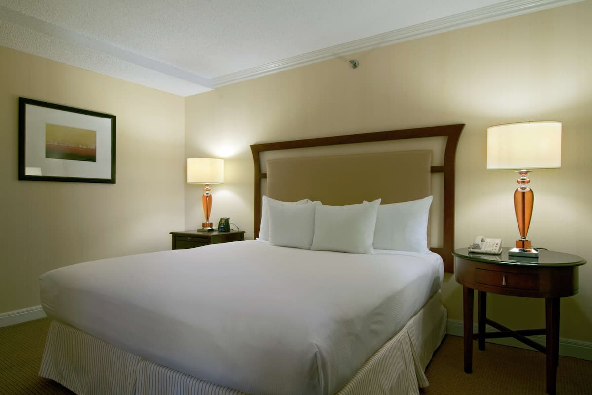 Quarto de hotel com uma cama grande, duas luminárias de cabeceira, um quadro na parede e uma mesinha com um telefone e uma luminária.