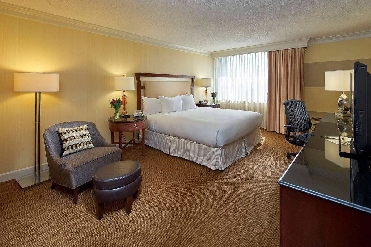 Quarto de hotel com uma cama grande, uma escrivaninha com um computador, uma cadeira, uma mesinha de cabeceira com abajur e um sofá.