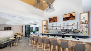 Uma área de bar moderna com um balcão comprido, banquetas e um espaço de estar com sofás e mesas.