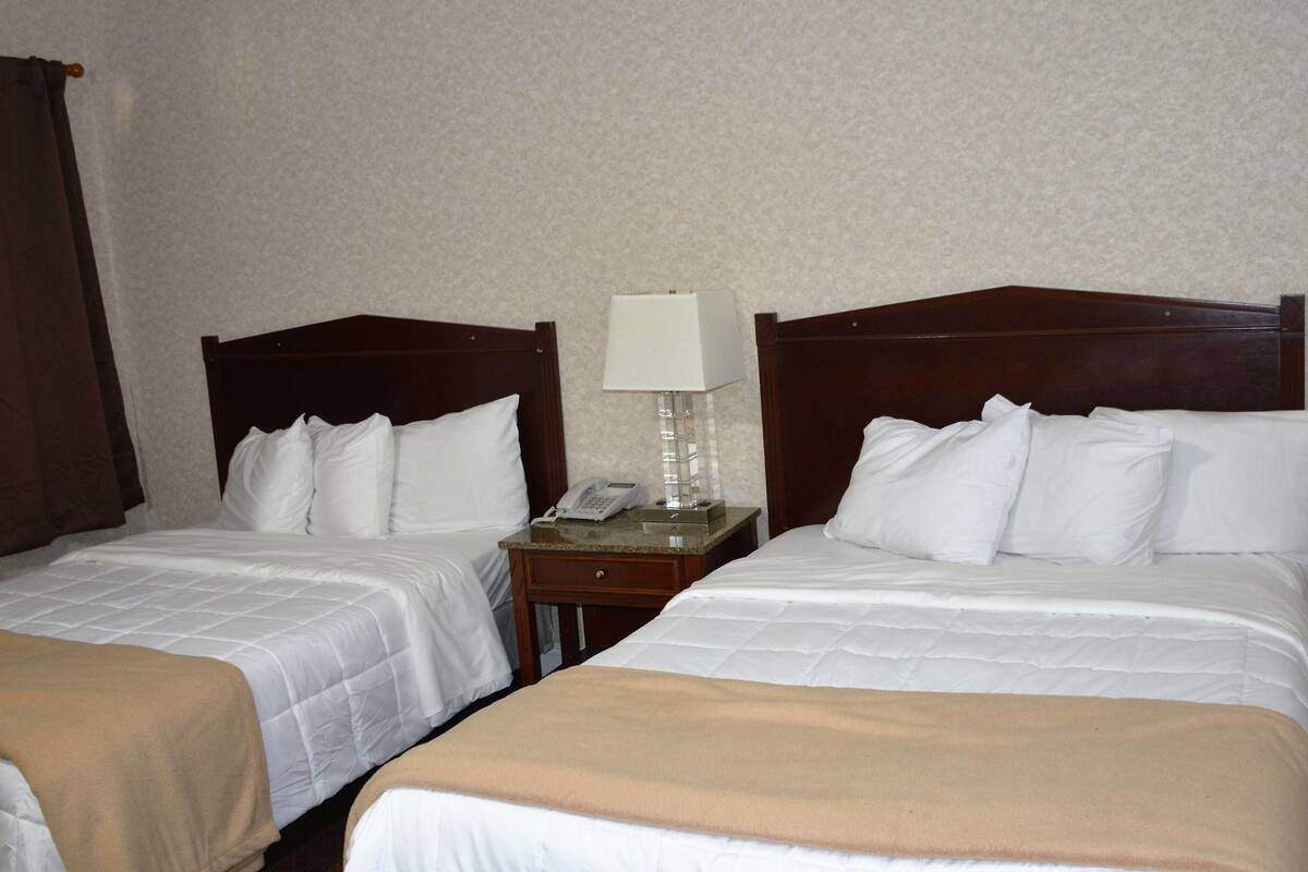 Quarto de hotel com duas camas de solteiro, cada uma com roupa de cama branca e cabeceira de madeira. Há uma mesinha de cabeceira entre as camas, com uma luminária e um telefone.