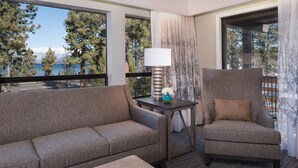 Quarto de hotel com sofá, poltrona e mesa com abajur. Vê-se árvores e montanhas pelas janelas.