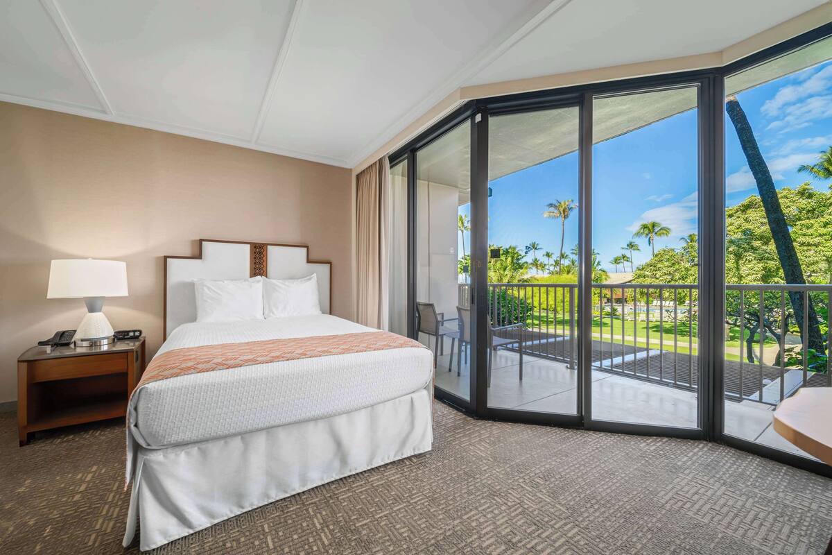 Quarto de hotel com cama, mesa de cabeceira com luminária, vista para uma varanda com mobiliário e céu limpo.