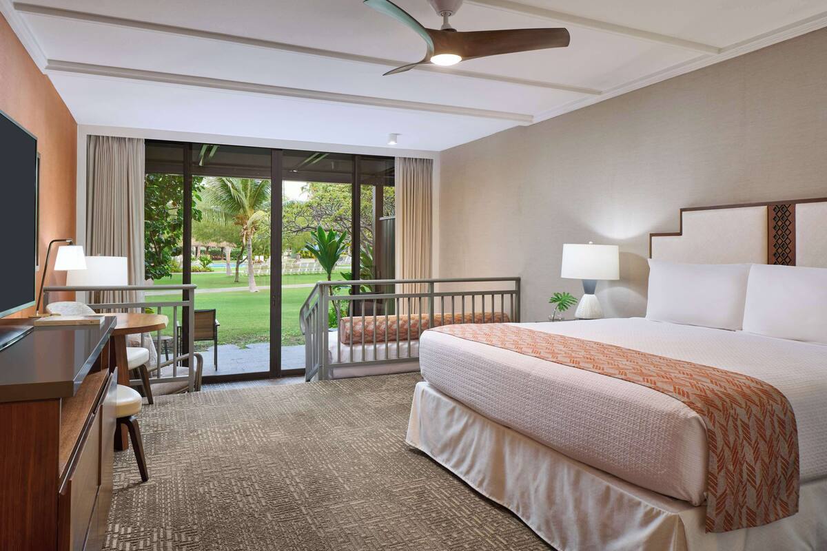 Quarto de hotel com cama, escrivaninha com cadeira, varanda com vista para áreas verdes e ventilador de teto.