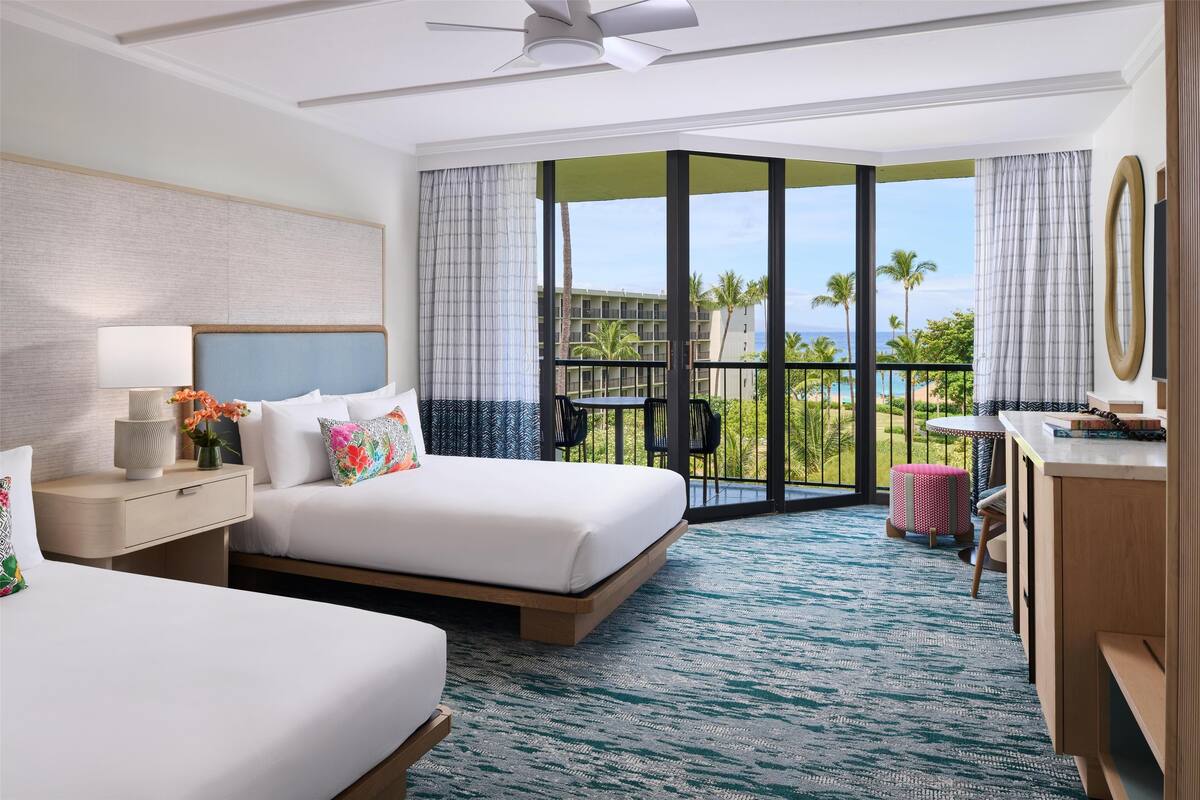 Quarto de hotel com duas camas, uma escrivaninha, uma cadeira e varanda com vista para a praia e palmeiras.