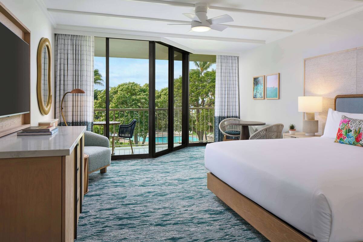 Quarto de hotel com cama, escrivaninha, cadeira, varanda e vista para árvores, e ventoinha no teto.
